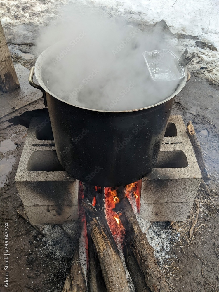 Maple sap boiling in a large pot over a fire to evaporate down into ...