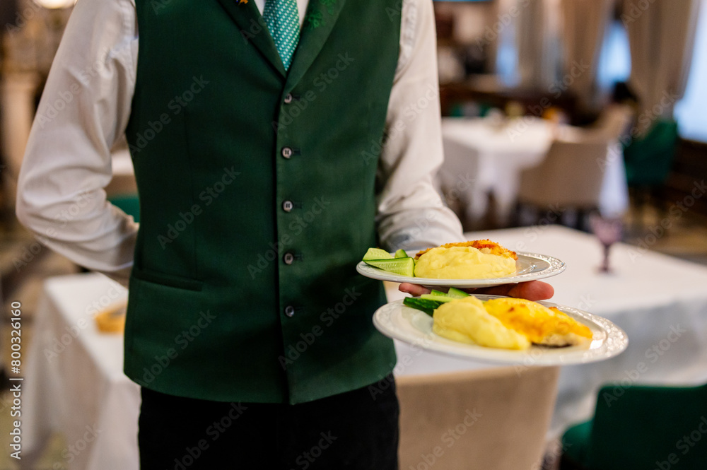 waiter in a green vest and tie serves two plates of delicious food in ...