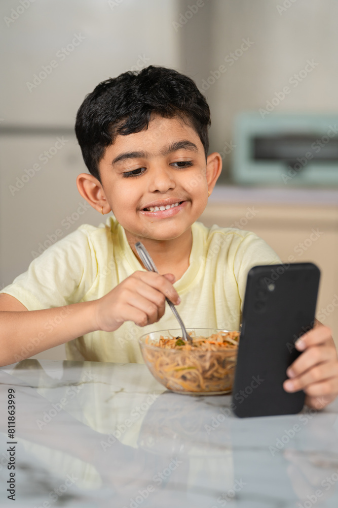 vertical shot of Indian preteen kid eating noodles while using mobile ...
