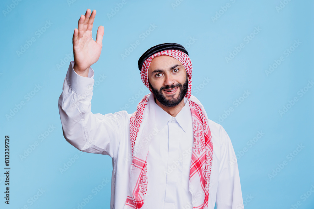 Smiling muslim man dressed in traditional clothes raising hand studio ...