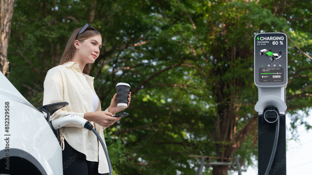 Young woman with coffee cup and sustainable urban commute with EV electric car recharging at ...
