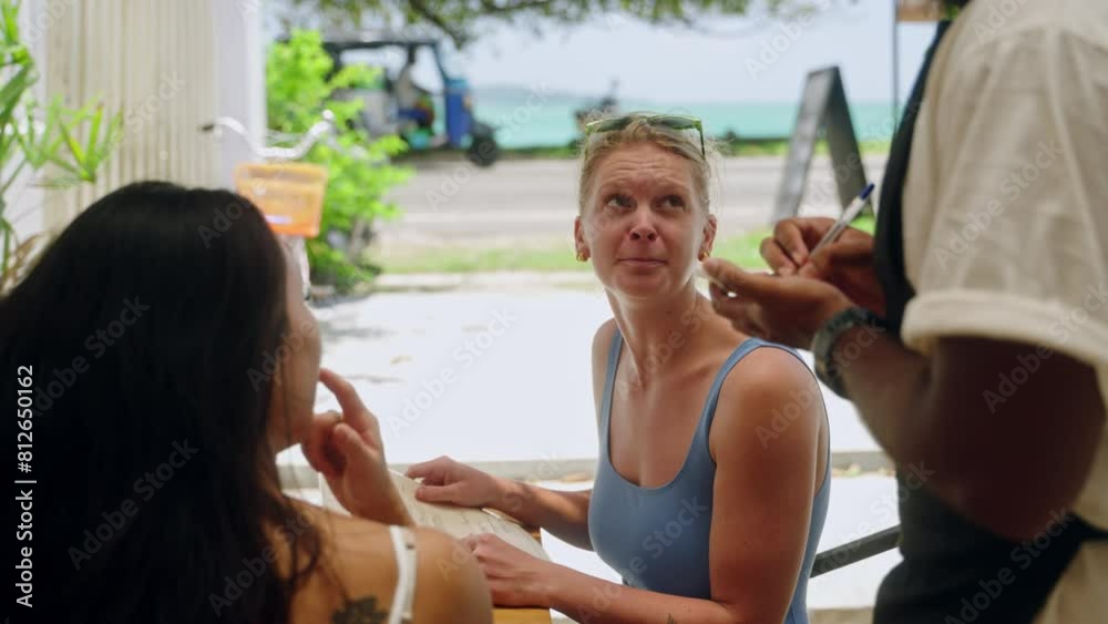 Multiethnic women seated at tropical cafe, engaging waiter taking order ...