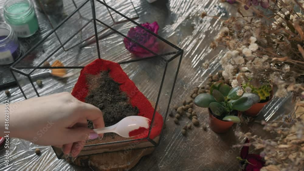 A woman pours multicolored earth into a florarium in her workshop. At ...