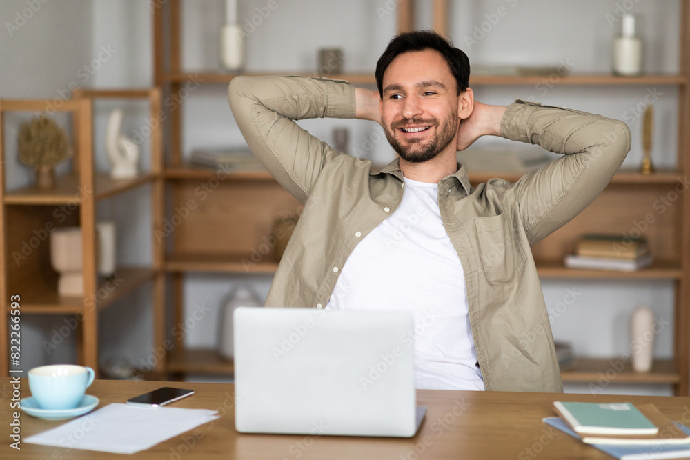 A young man with a cheerful expression sits back at his desk in a well ...