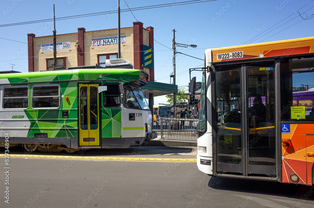 A tram is at a tram stop with a bus passing by in the same street but ...