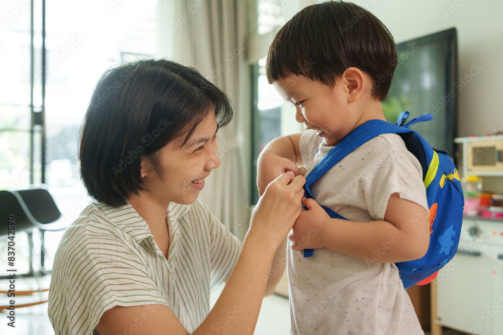 Smiling Asian mother adjusts blue backpack on happy young son. Child laughs while mother ensures ...