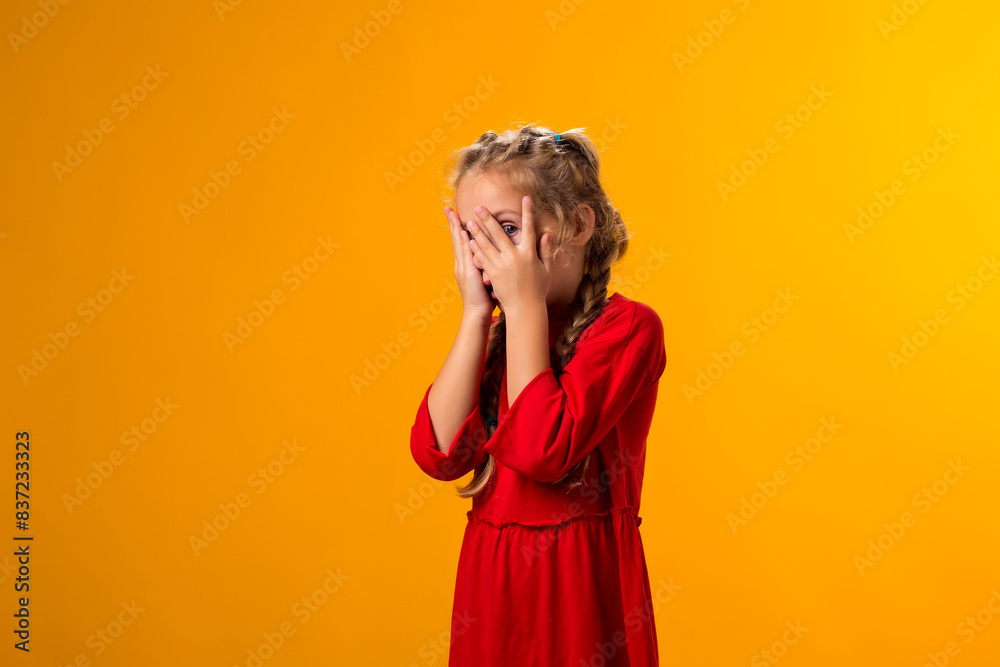 Scared little girl covers her face with her hand over yellow background ...