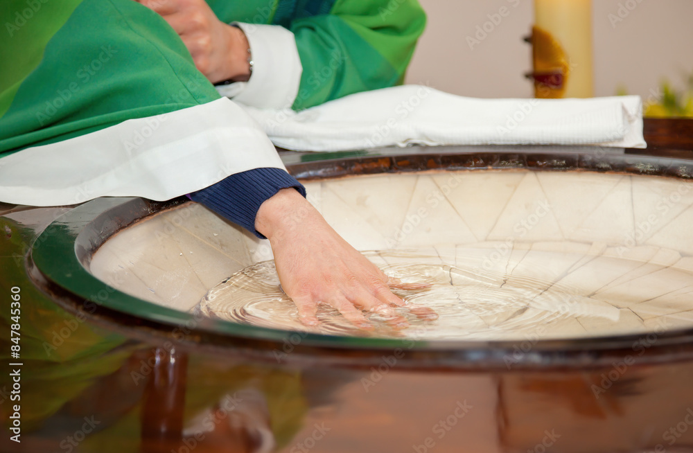 Priest in green liturgical vestments blessing holy water in a marble baptismal font inside a ...
