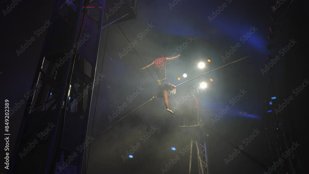An acrobat jumps over a man on a tightrope under the dome of the circus ...