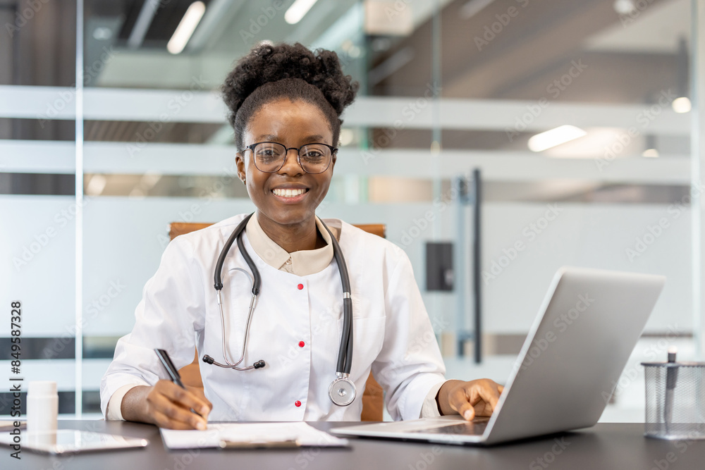 Smiling doctor working at desk with laptop, writing notes in a modern ...