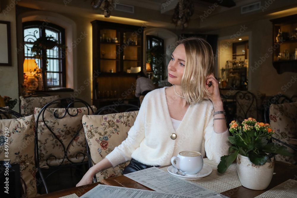 Blonde woman in white jacket sits inside in the cozy cafe with a cup of ...