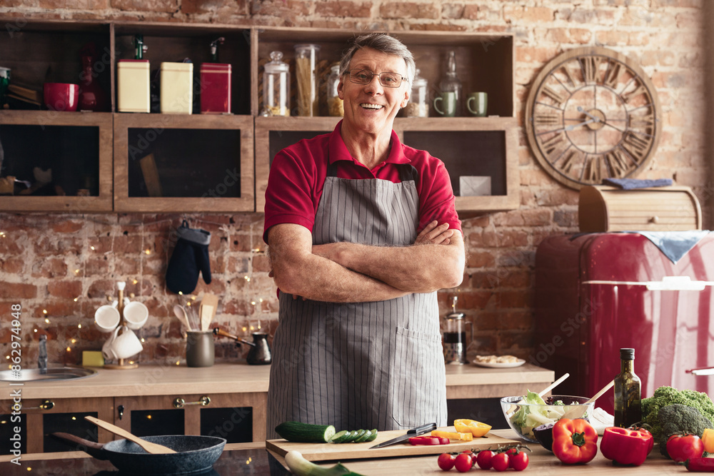 An older man stands in his kitchen, arms crossed, wearing a gray apron ...