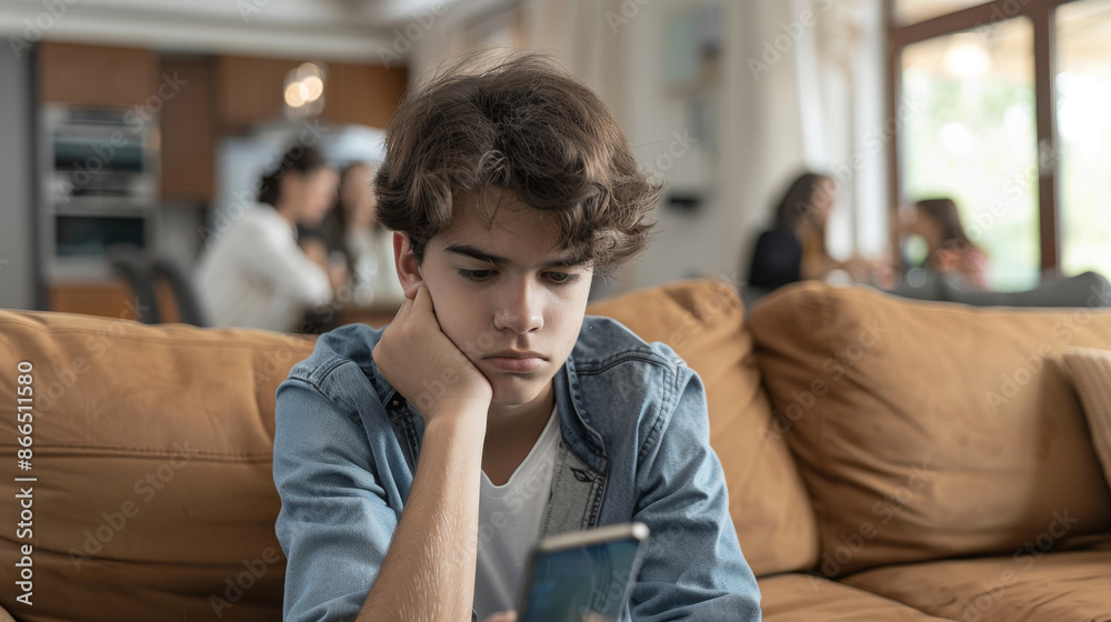 Sad teenage boy scrolling his phone, Behind him, happy family is eating ...