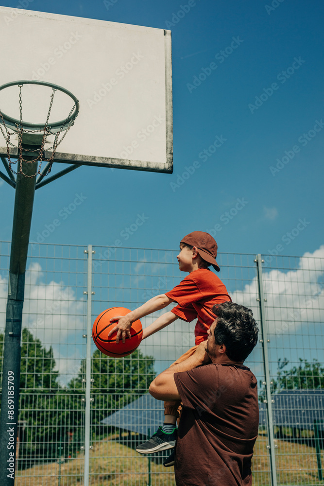 Boy throwing basketball in hoop and sitting on father's shoulder at ...