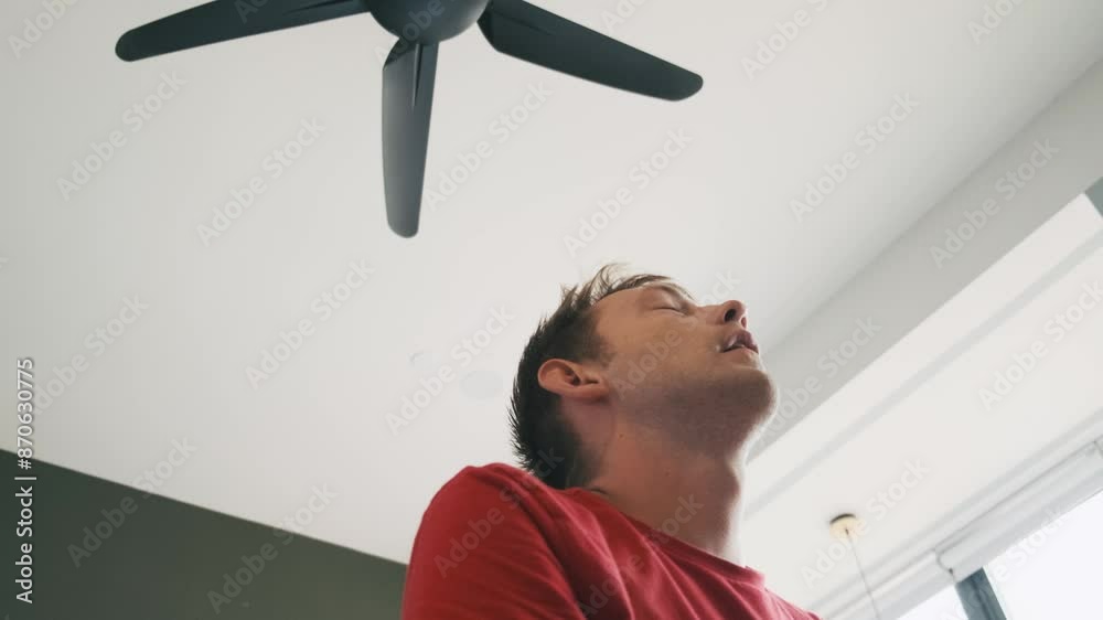Exhausted man sitting under ceiling fan at home, cooling off during hot ...