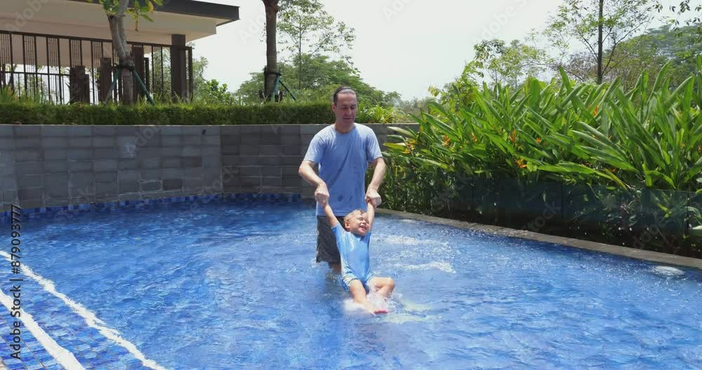 Father stands in shallow pool, holding his baby son's hands. He raises ...