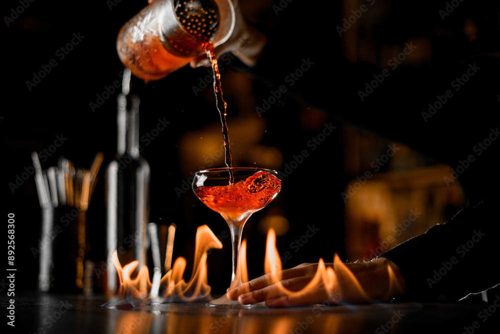 Bartender pours drink into chilled glass amidst a burning bar counter. Stock Photo | Adobe Stock