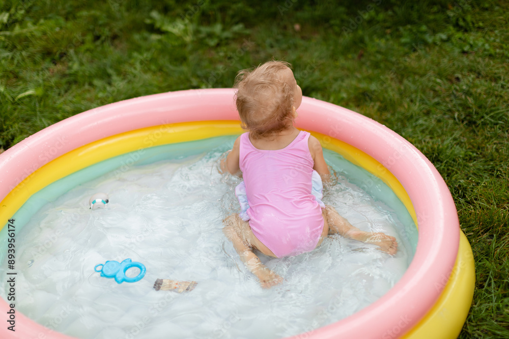 Baby in a pink swimsuit is swimming in inflatable pool in the backyard ...