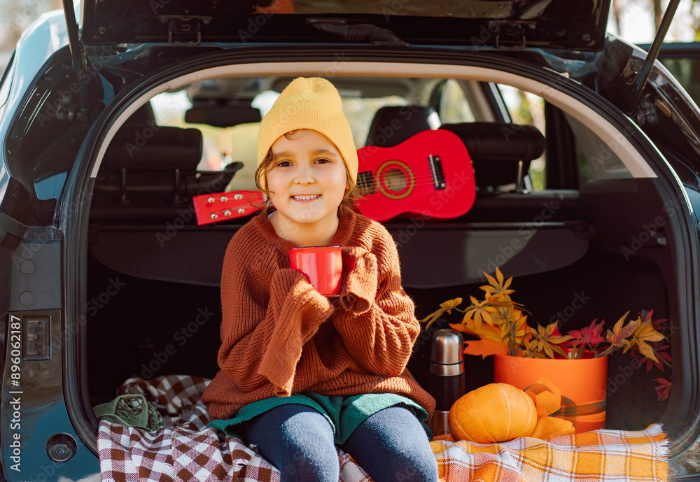 Little cute smiling girl sitting in open car trunk and drinking cocoa ...