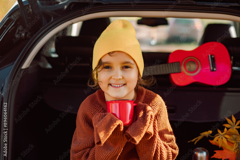 Little cute smiling girl sitting in open car trunk and drinking cocoa ...