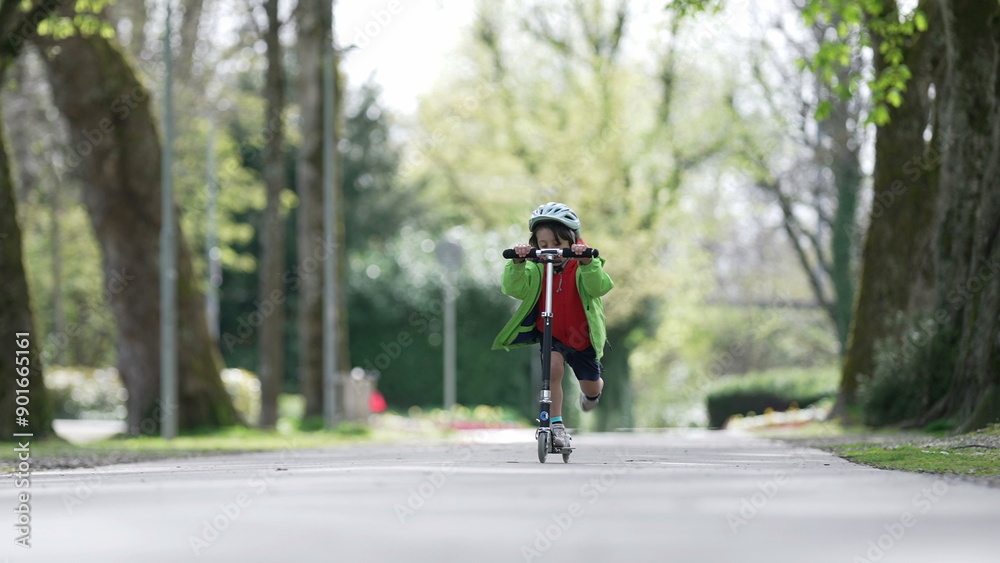 Young child riding a scooter on park path, wearing green jacket and ...