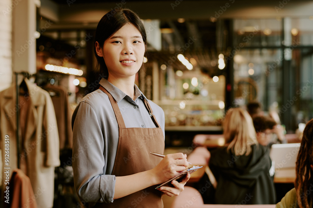 Smiling Asian waitress in apron holding notepad in busy cafe, engaging ...