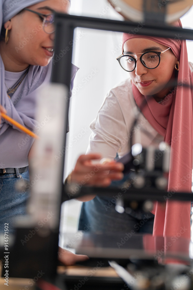 Two women in hijabs are seen adjusting the settings on a 3D printer ...