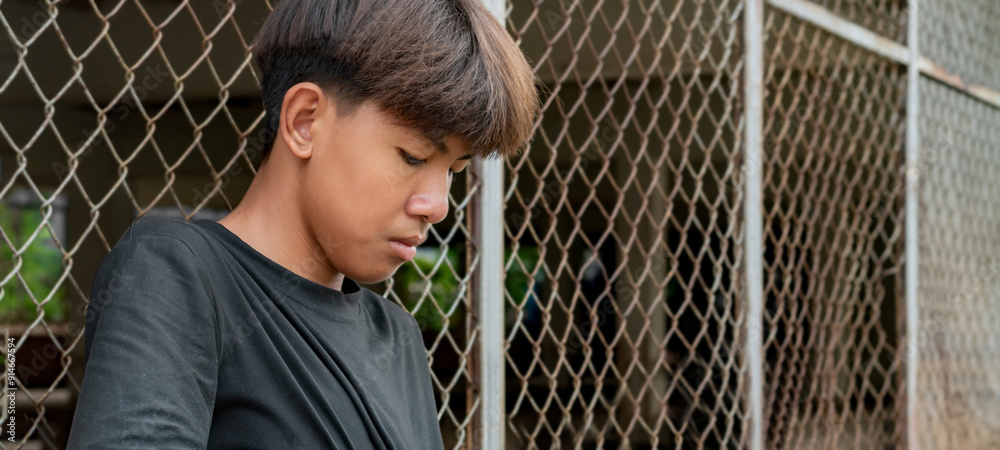 Asian teenboy in a black shirt sits pressed against a metal fence panel ...