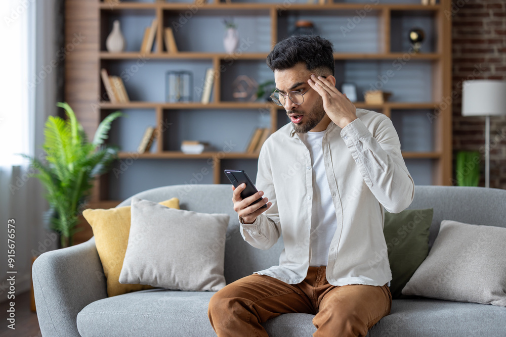 Confused young Indian man is at home, sitting on the sofa, looking ...