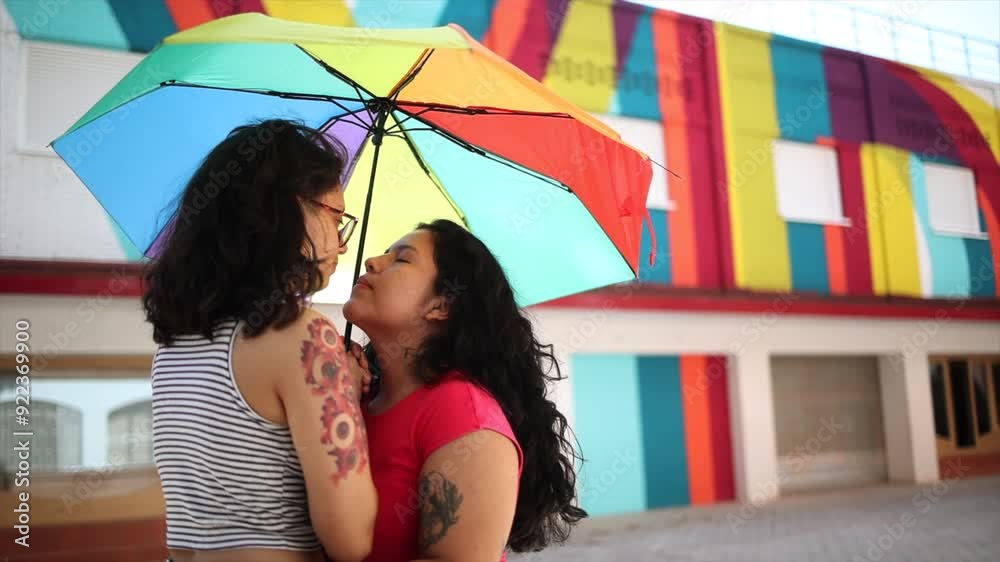 Lesbian couple sharing a kiss under a rainbow umbrella, celebrating love and pride against a ...