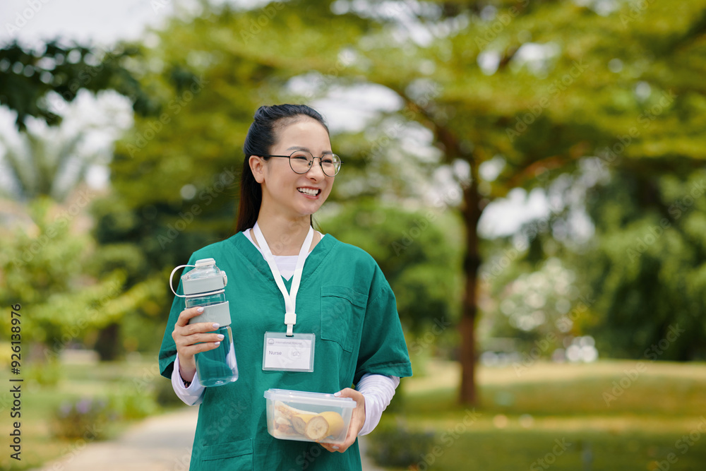 Nurse smiling while holding water bottle and lunch box in outdoor ...