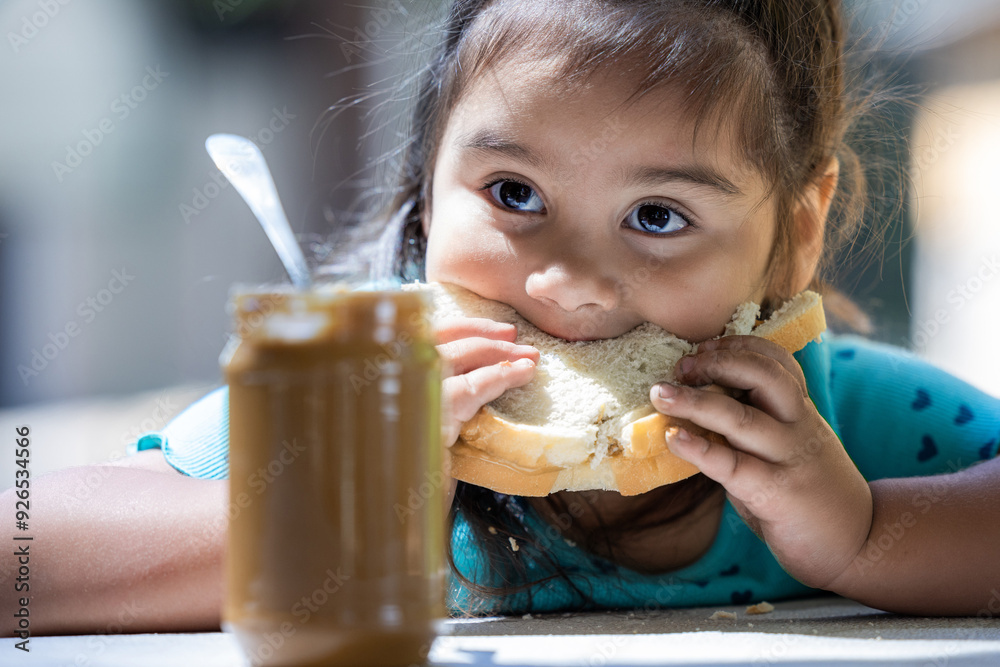 A young girl is eating a sandwich with a jar of peanut butter next to ...