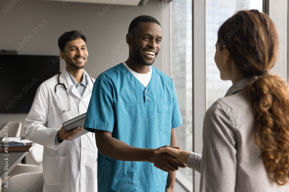 Female shake hands with young smiling African doctor in blue scrubs, as ...