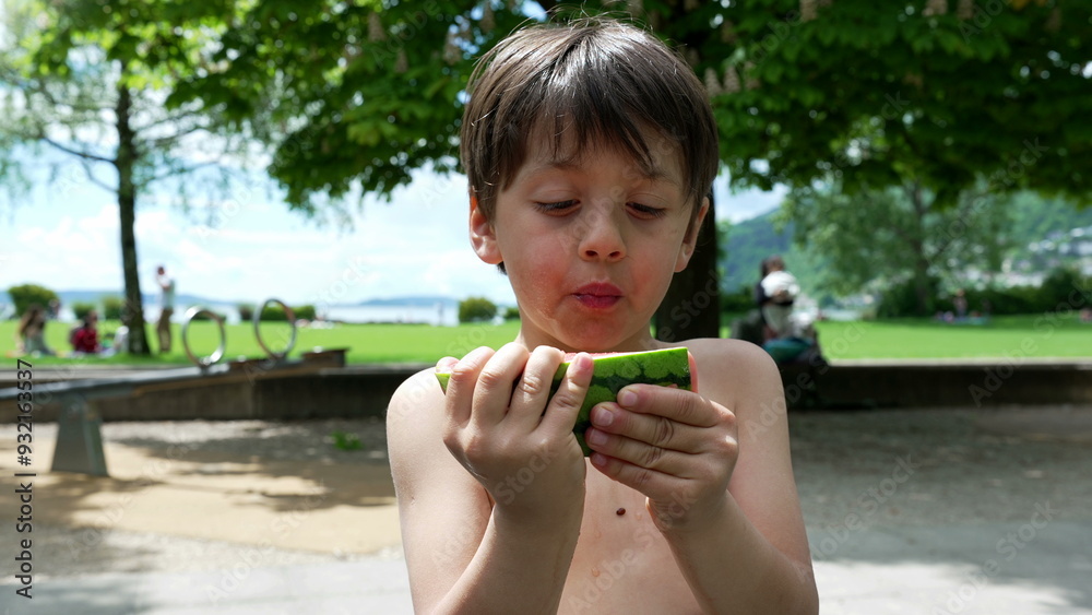 Young boy eating watermelon, standing shirtless in a park, enjoying a ...