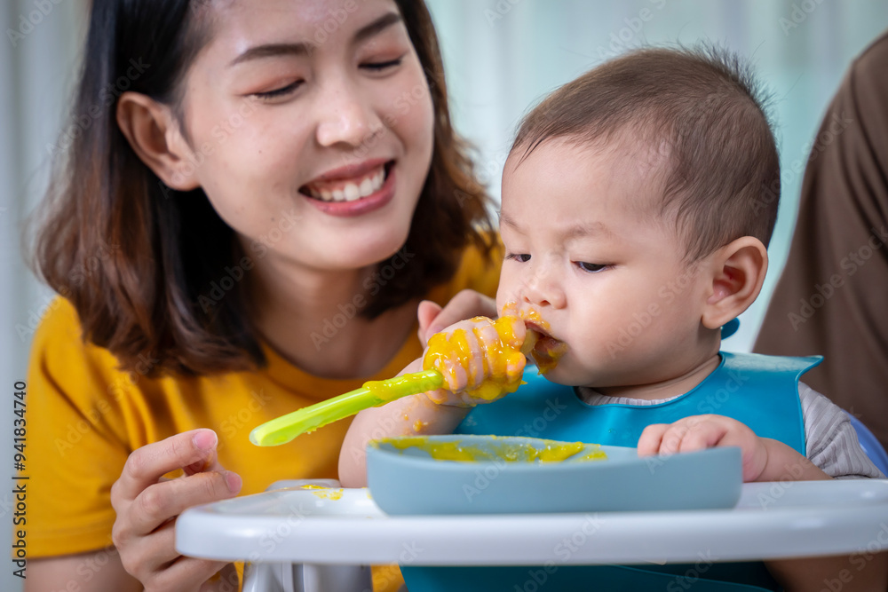 A woman feeds a baby in a high chair. The baby is wearing a bib and is ...