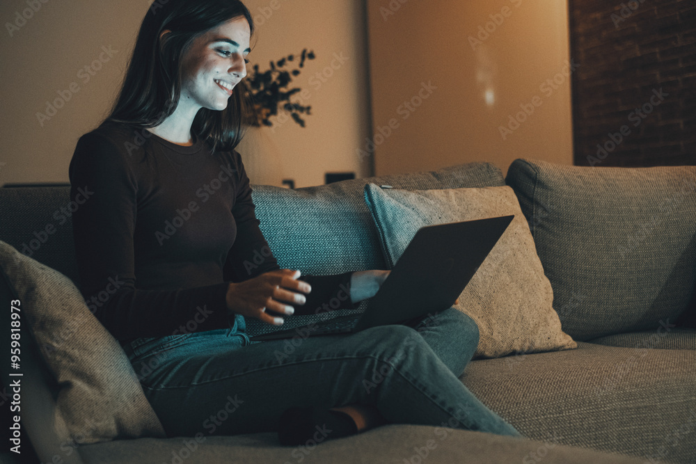 Young freelance woman working late into the night on a laptop, comfortably seated on a sofa in ...