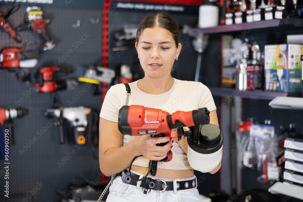 Woman customer examines electric spray gun airbrush in tools department of store. Girl consider ...
