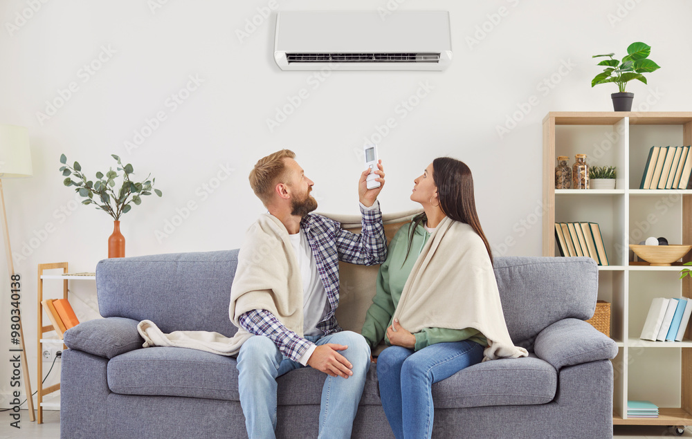 Young frozen couple man and woman turning on air conditioner sitting on ...