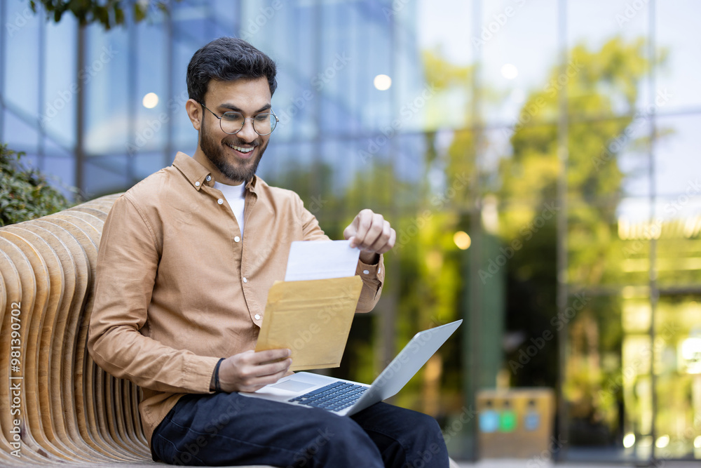 Smiling man sitting on bench reading letter from envelope with laptop ...