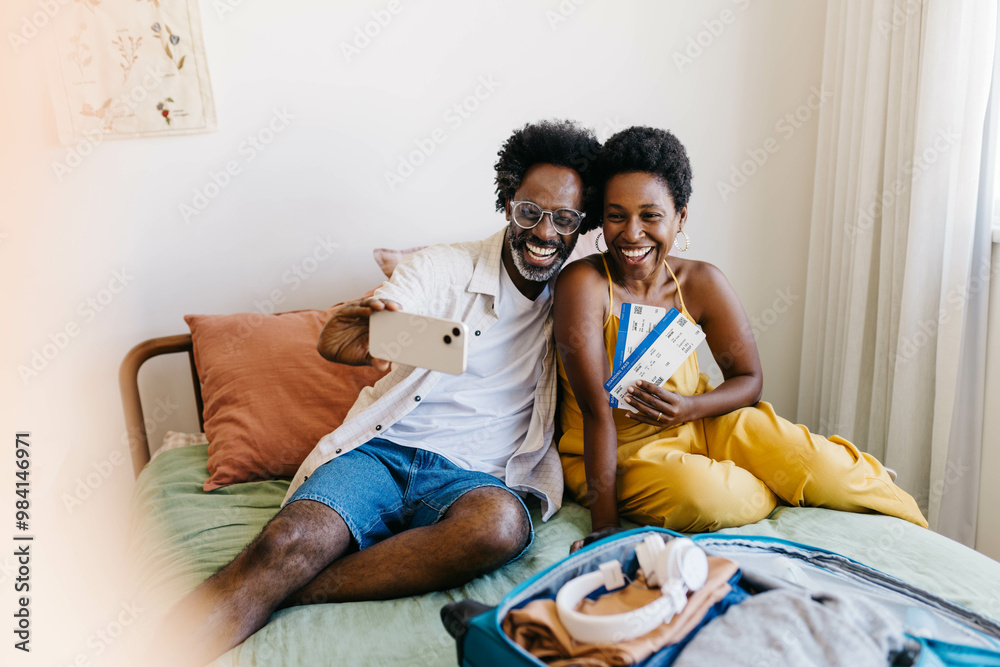 Excited couple smiling on bed with flight tickets, making a video call ...
