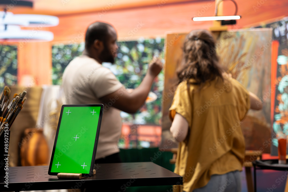 Two artists painting on a canvas next to green screen on tablet display ...