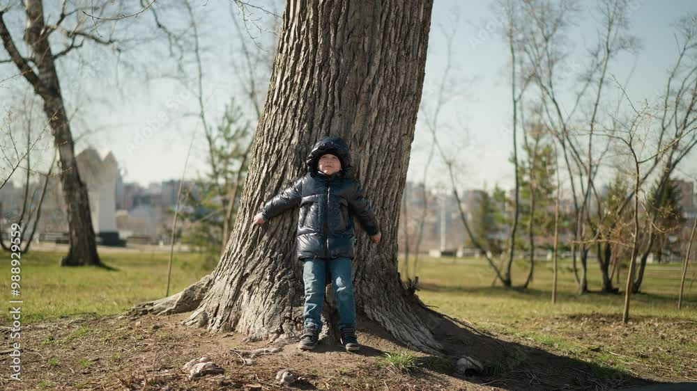A young boy leans his back against a large tree trunk, standing with ...