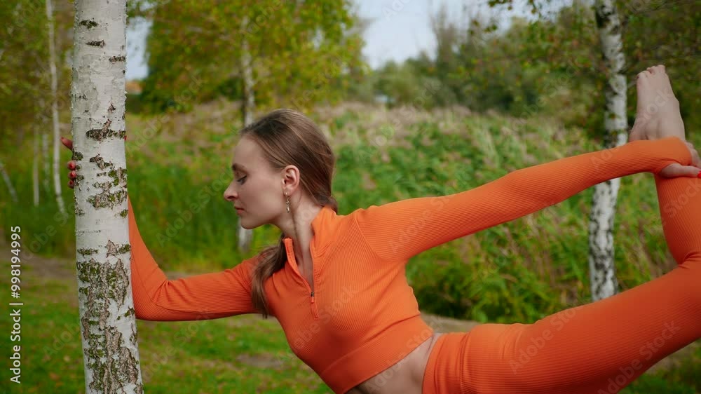 A woman in a vibrant orange outfit stretching in a park during her yoga ...