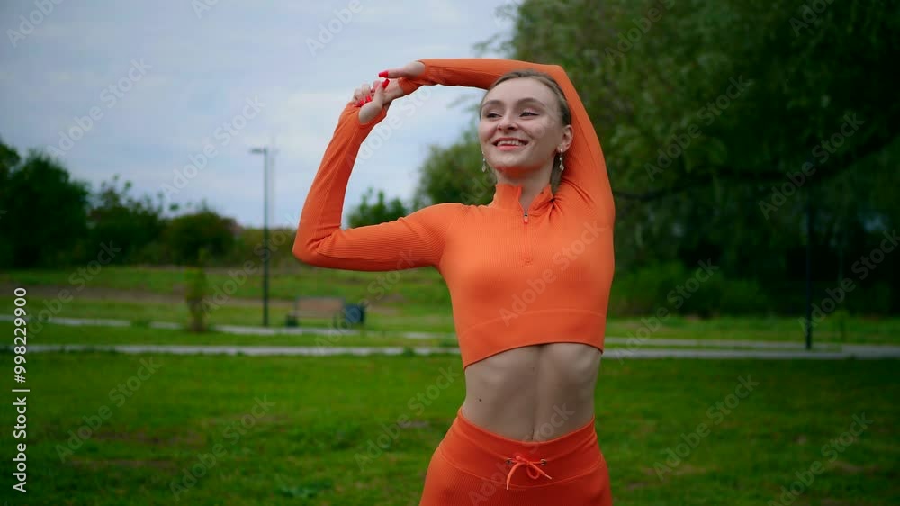 A woman in a vibrant orange outfit stretching in a park during her yoga ...