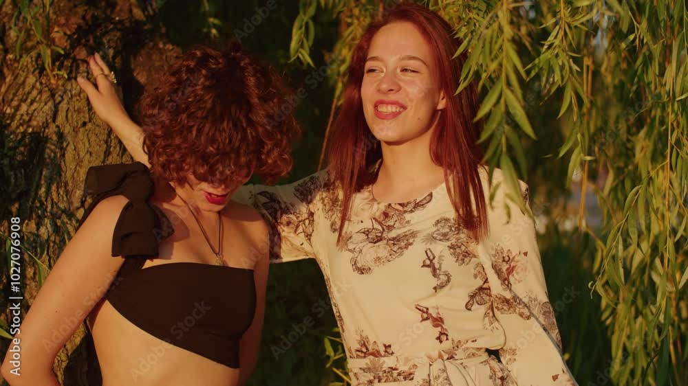 Two women laughing together near a tree in warm sunlight. Outdoor ...