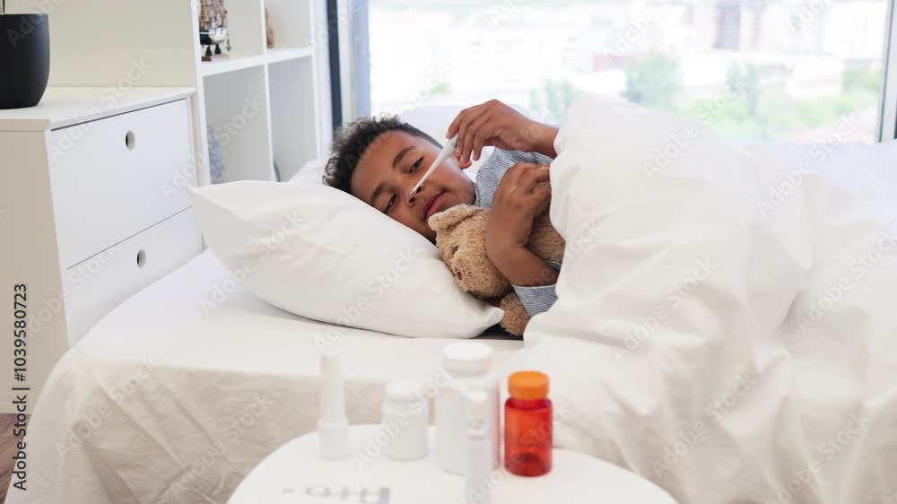 African boy sick with flu lies in bed measuring temperature with thermometer. Young child holds ...