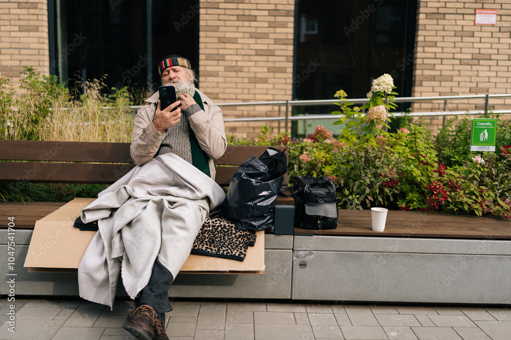 Homeless elderly man sitting on urban bench with blanket, using ...