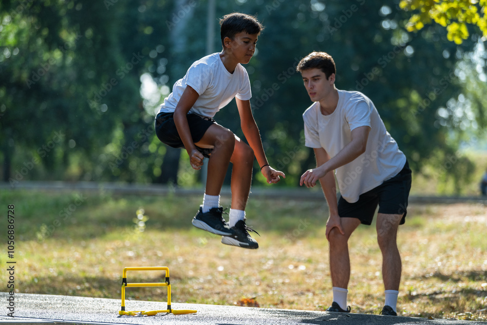 Young boy practices jumping hurdles with a trainer outdoors, promoting ...