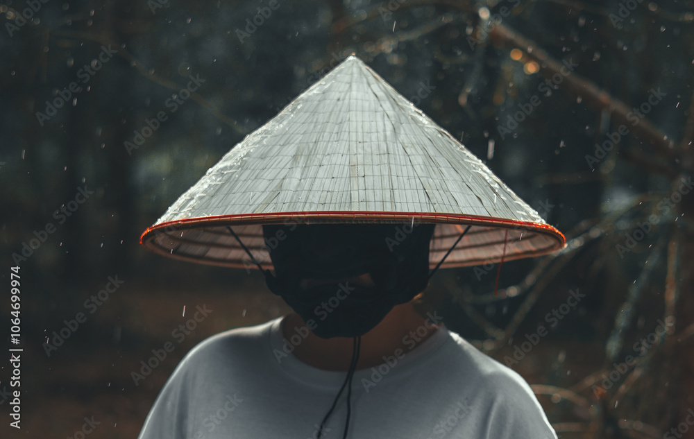 A man wearing a traditional conical hat and mask stands in the rain ...