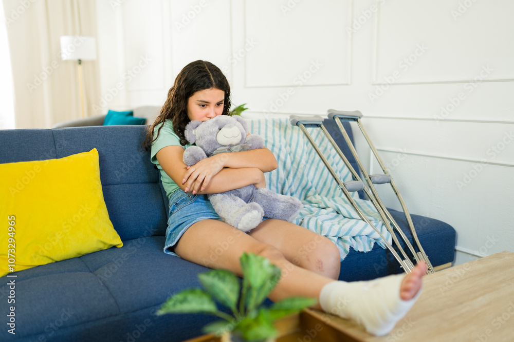 Sad teen girl with broken leg and crutches hugging teddy bear at home ...